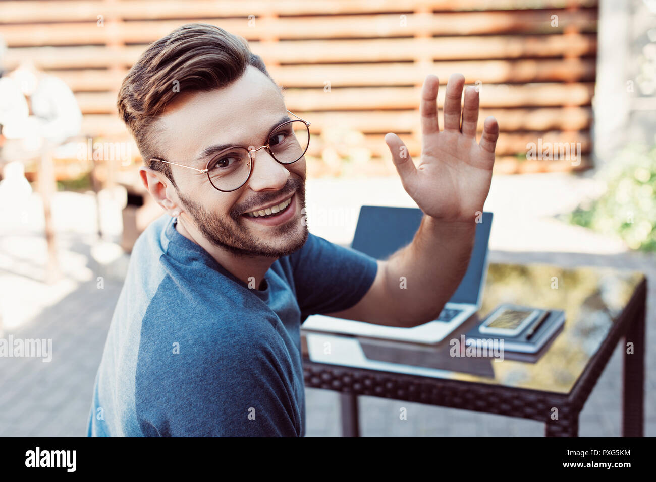 smiling handsome man waving hand and looking at camera at street cafe ...