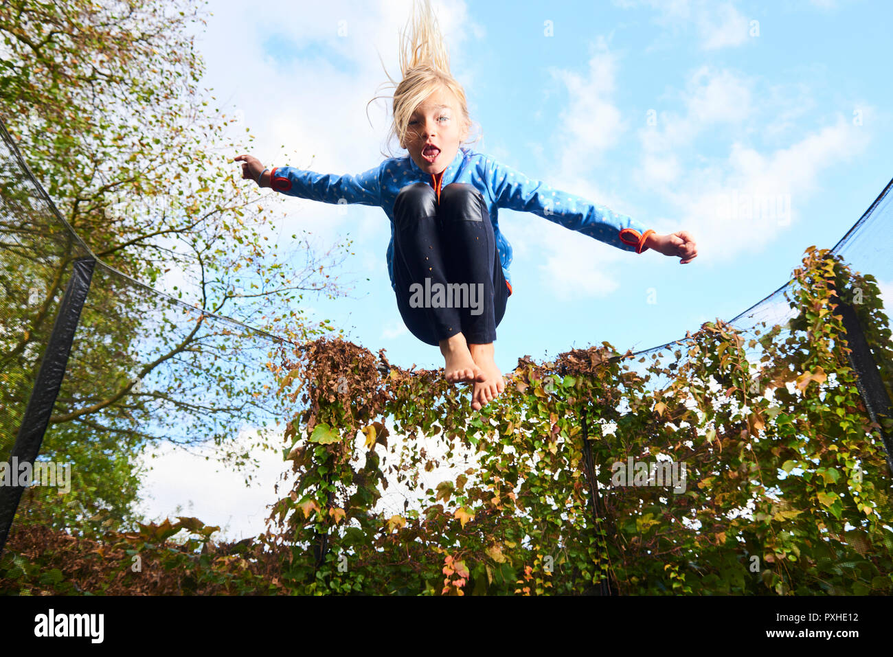 Child cute blond girl playing and jumping on trampoline with greenery ...