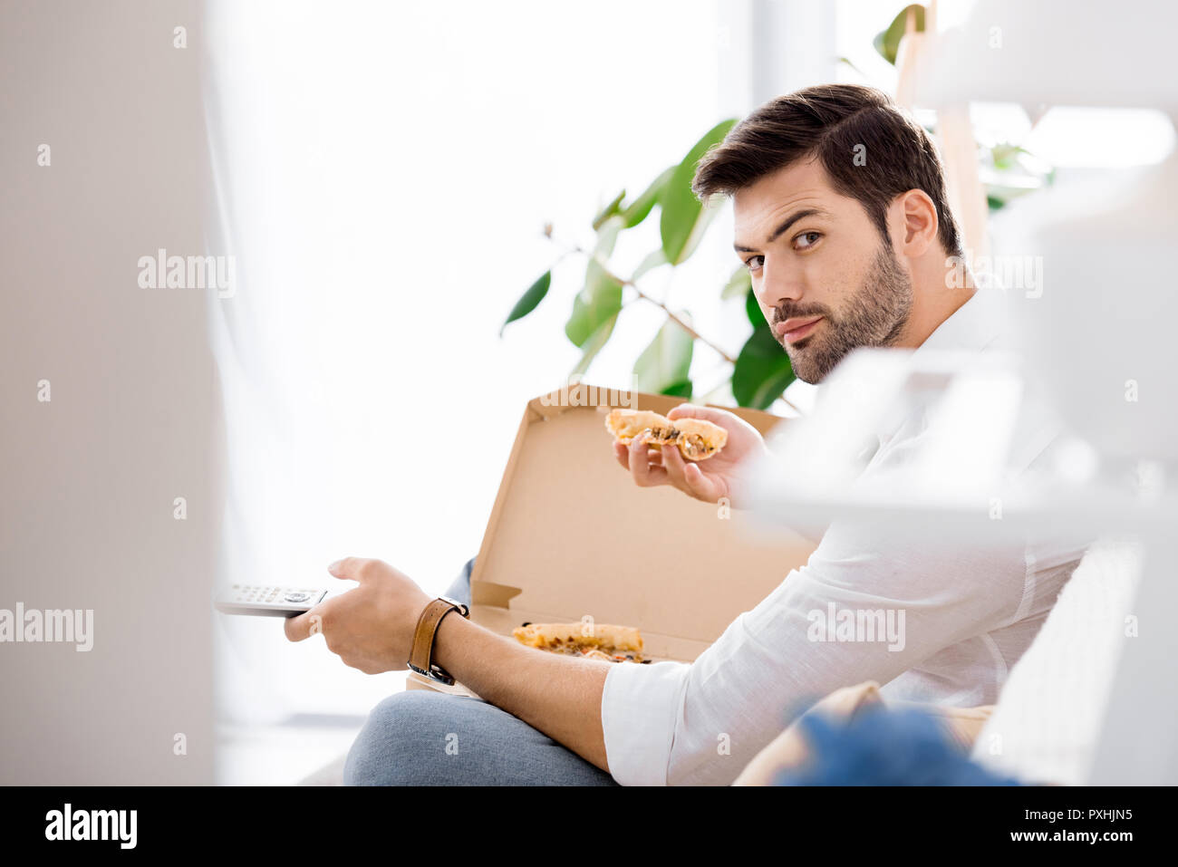 side view of young man with remote control in hand eating pizza alone ...
