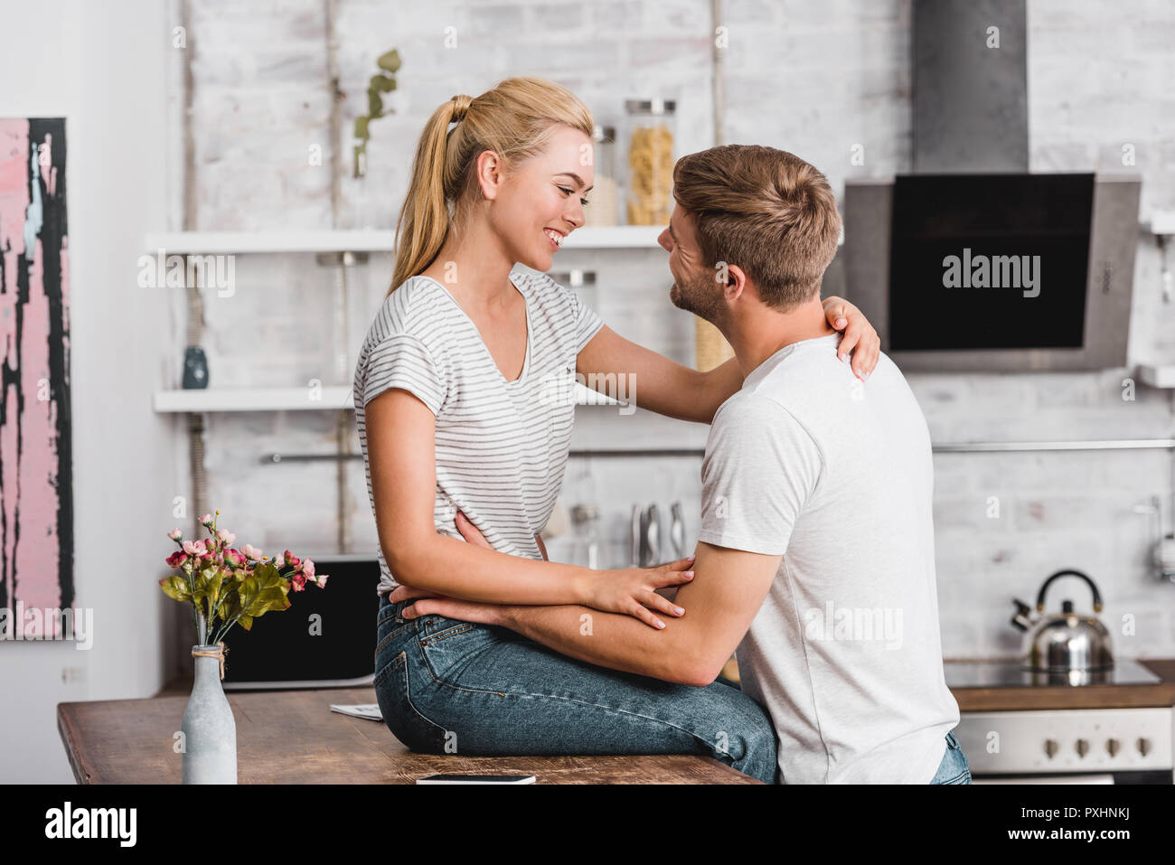 side view of happy girlfriend sitting on kitchen counter and looking at ...