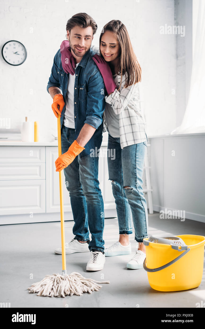 boyfriend cleaning floor in kitchen with mop and girlfriend hugging him ...