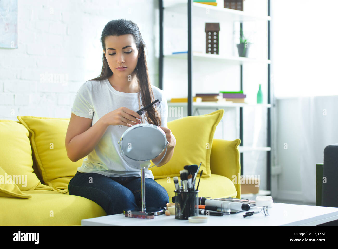 attractive woman brushing hair with comb while sitting on sofa at home ...