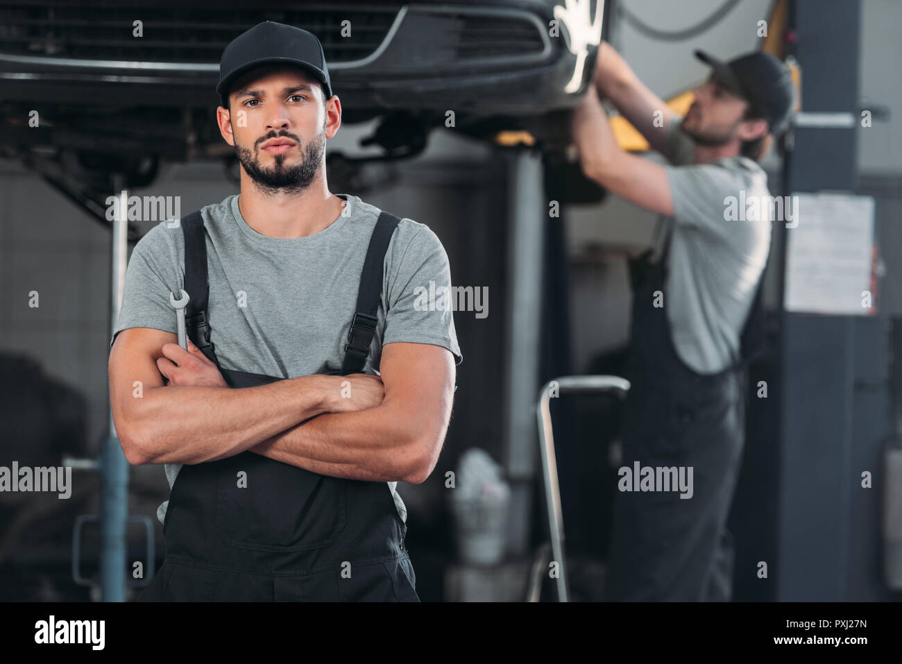 serious mechanic with crossed arms holding wrench, while colleague ...