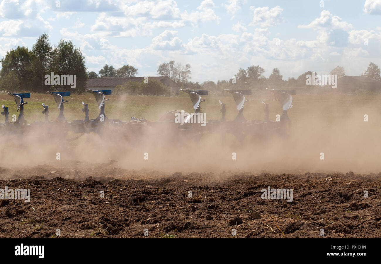 large blue plow plows the soil on the field, a lot of dust from the dry ...