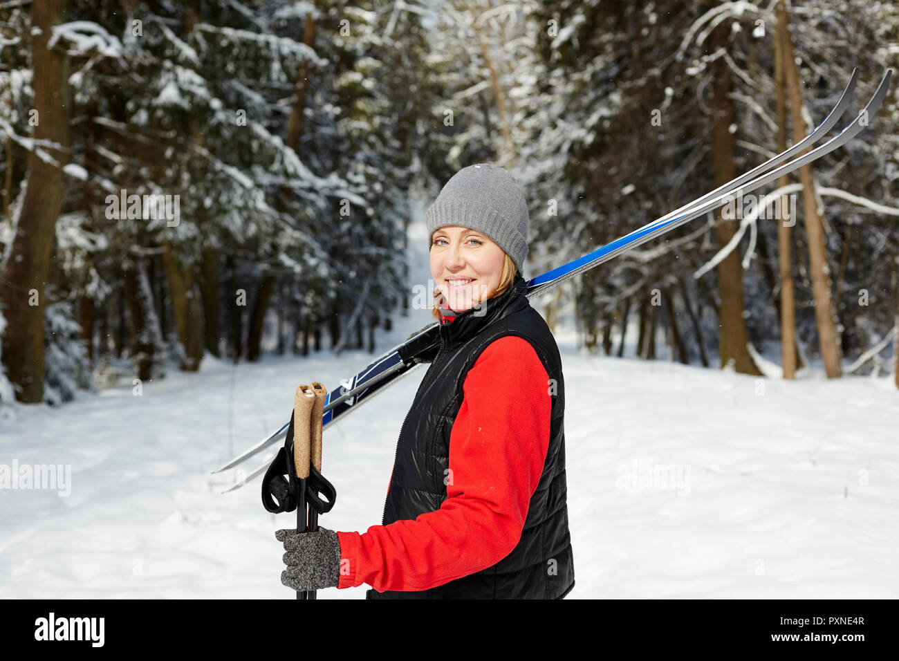 Happy mid-aged female in winter activewear carrying skiing equipment ...