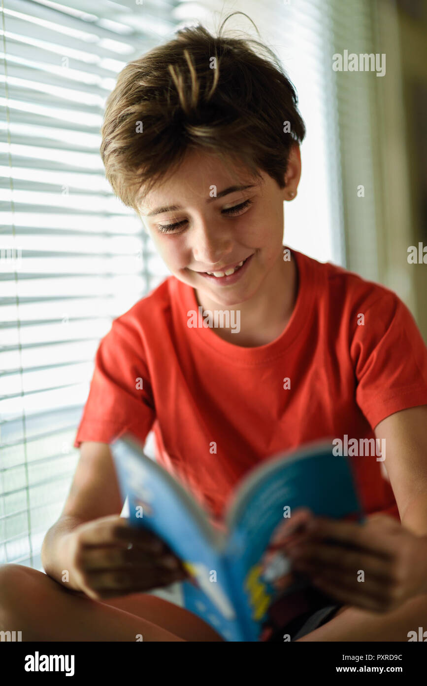 Portrait of happy little girl reading a book beside window at home ...