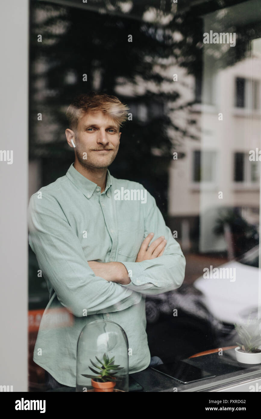 Young man standing at the window, with arms crossed, using ear buds ...