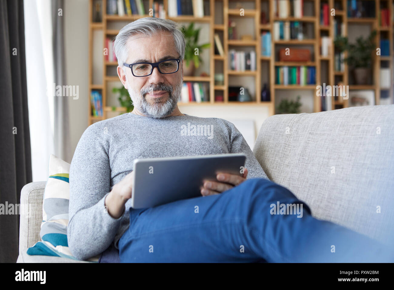 Portrait of mature man sitting on couch at his living room using tablet ...