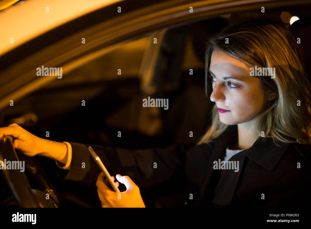 Pretty young woman using her smart phone while driving her car at night ...