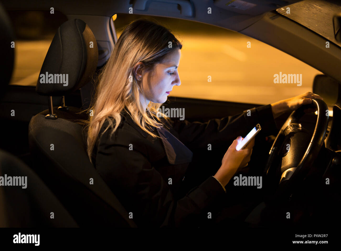 Pretty young woman using her smart phone while driving her car at night ...