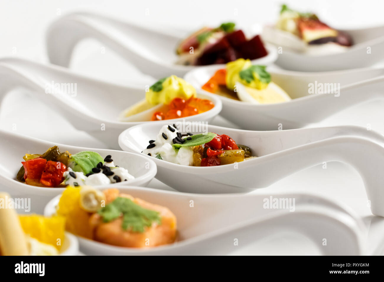 A row of mixed canapes in white ceramic spoons on white background ...