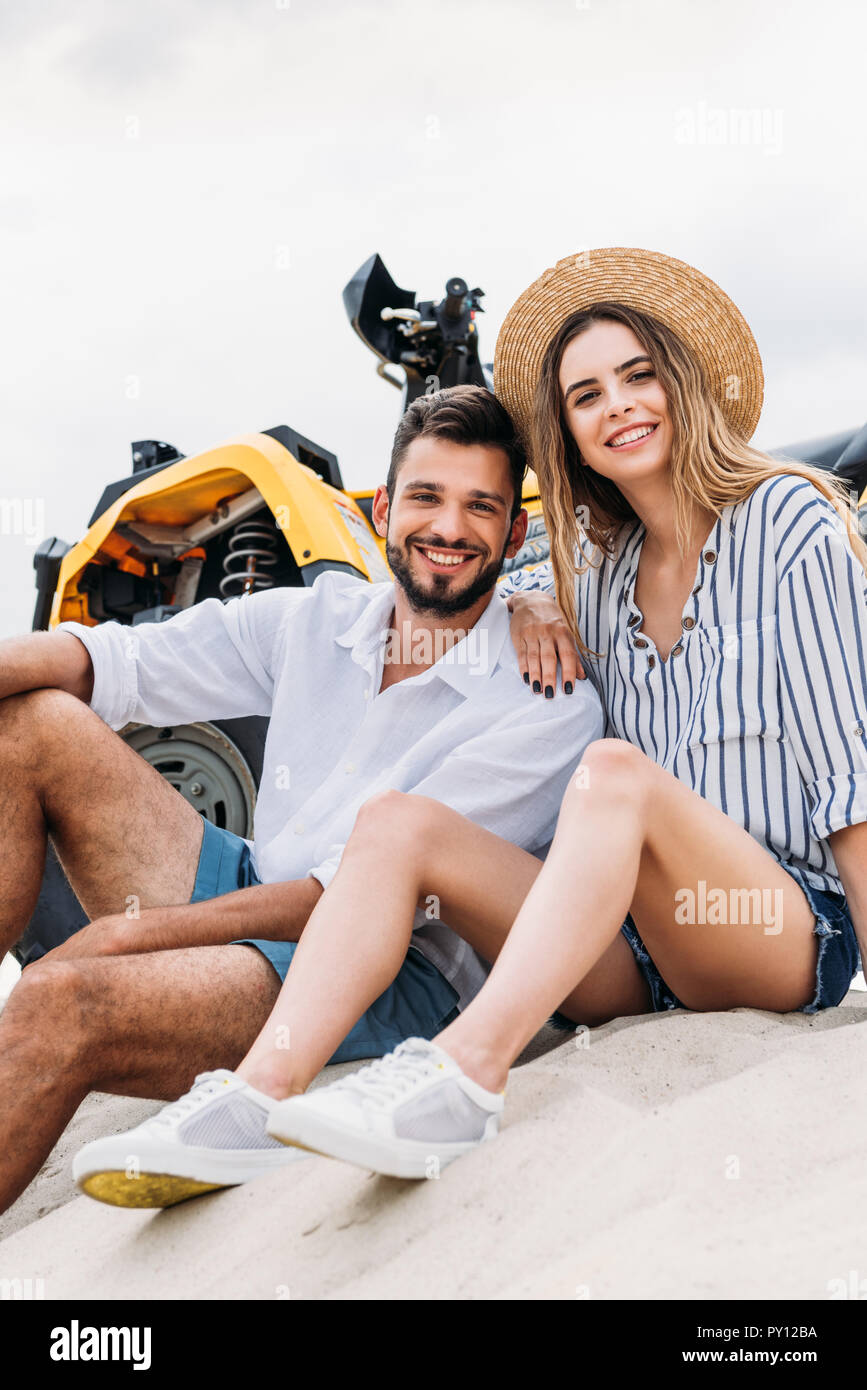 happy young couple sitting near ATV on sandy dune and looking at camera ...