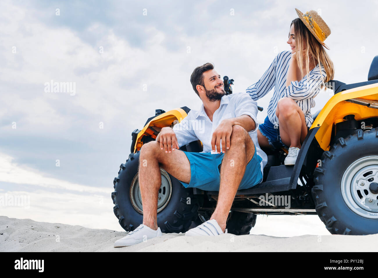 happy young couple sitting on ATV on sandy dune in front of cloudy sky ...