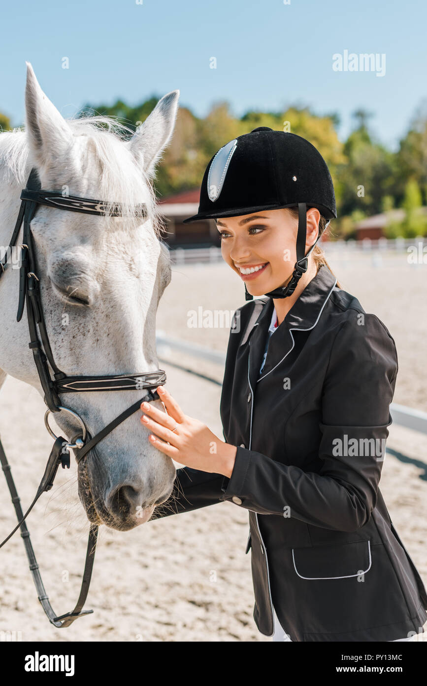 attractive smiling female equestrian standing near horse at horse club ...