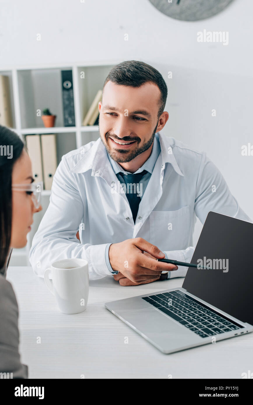 smiling doctor pointing on laptop with blank screen to patient in ...