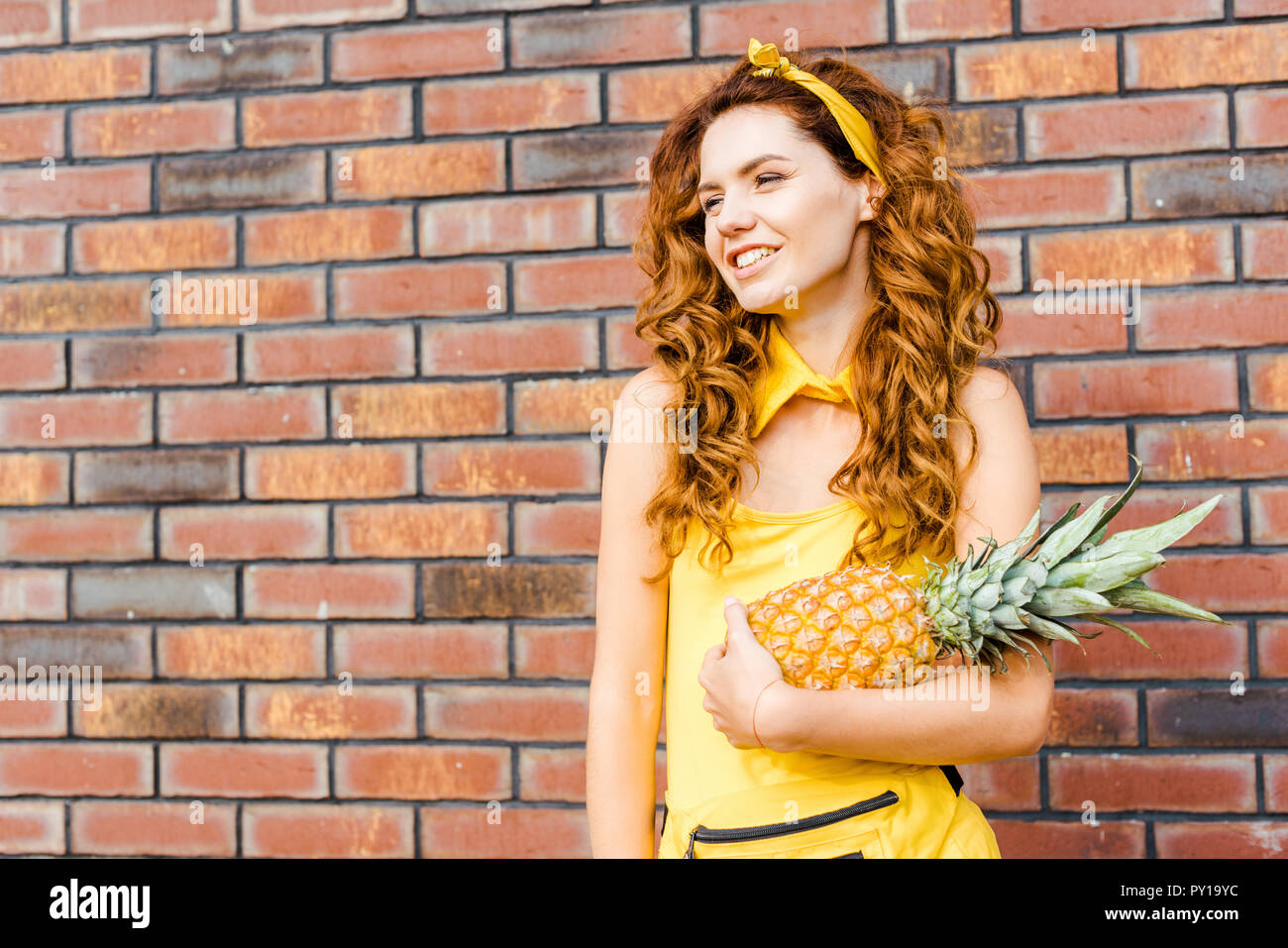 smiling young woman in yellow clothes holding pineapple in front of brick wall Stock Photo - Alamy
