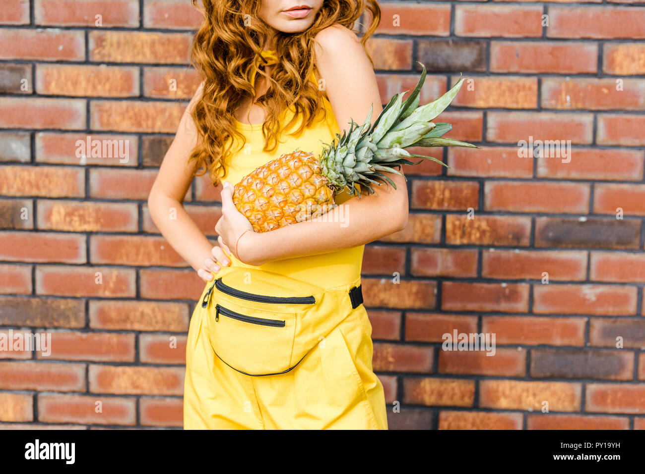 cropped shot of young woman in yellow clothes holding pineapple in front of brick wall Stock ...