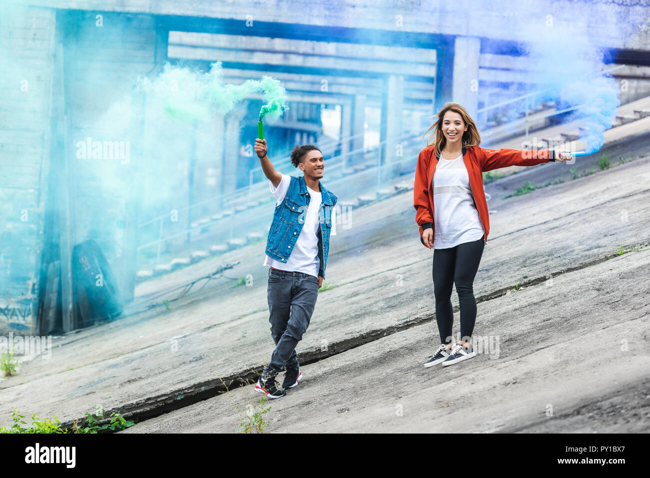 smiling multicultural couple holding colorful smoke bombs at urban ...