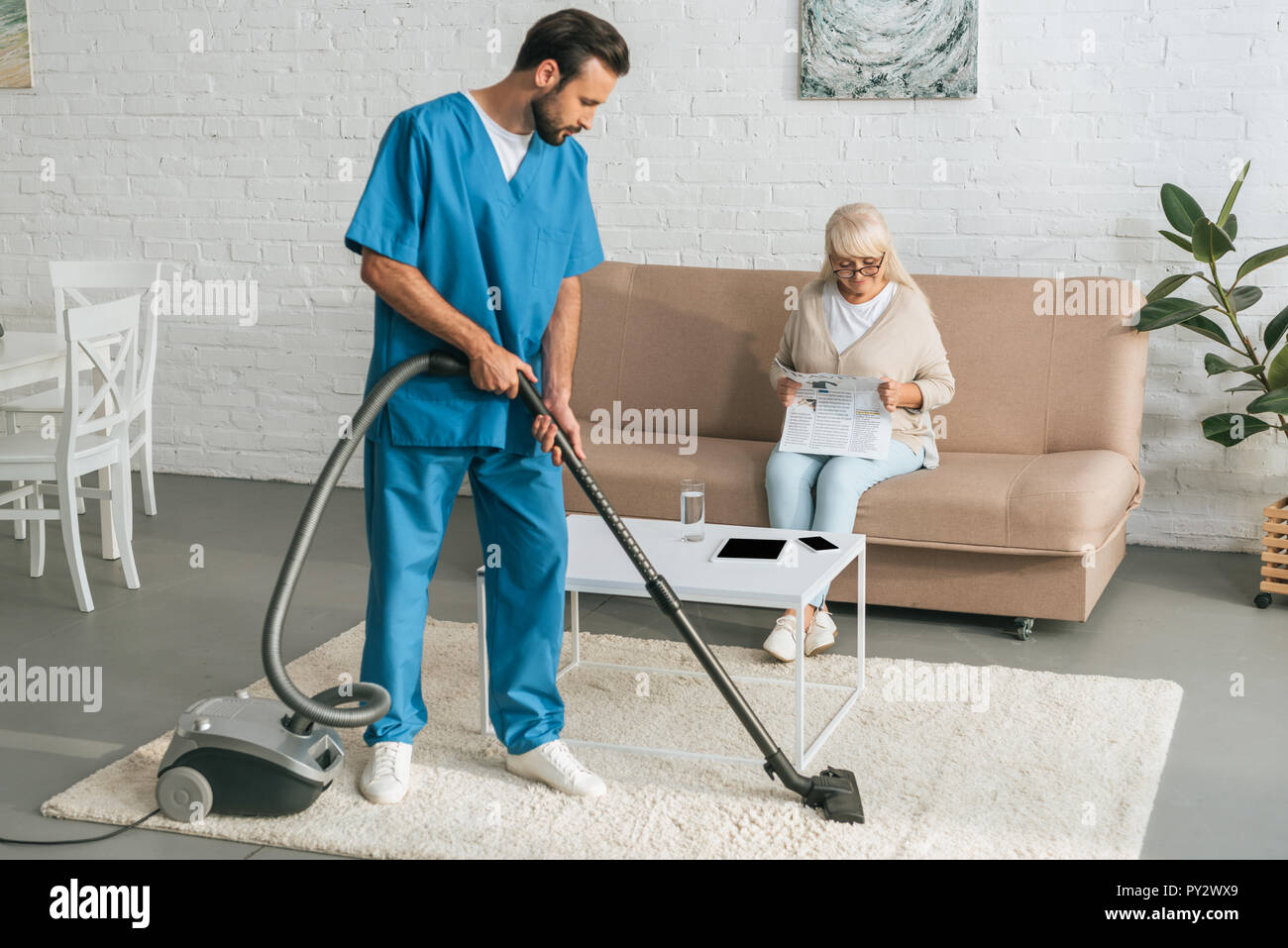 young social worker cleaning carpet with vacuum cleaner while senior ...