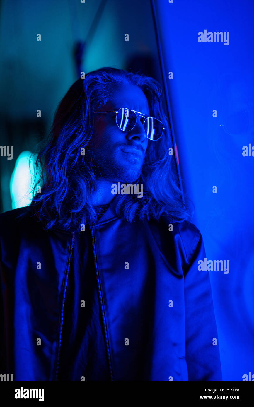 attractive young man in sunglasses under blue light on street at night ...