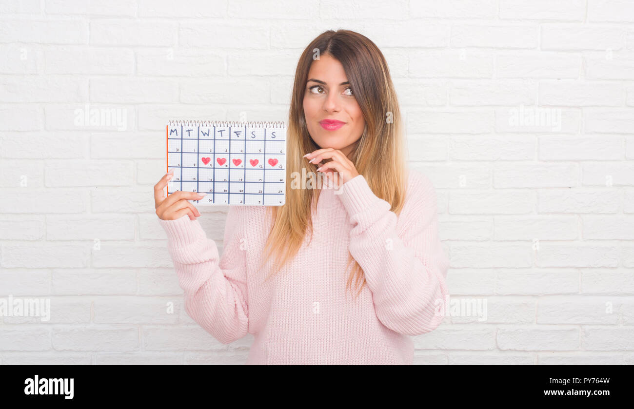 Young adult woman over white brick wall holding period calendar serious ...