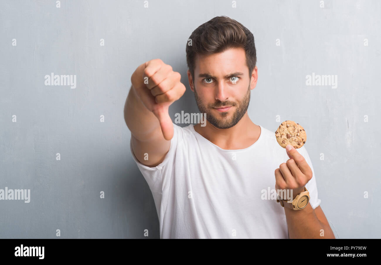Handsome young man over grey grunge wall eating chocolate chip cooky ...