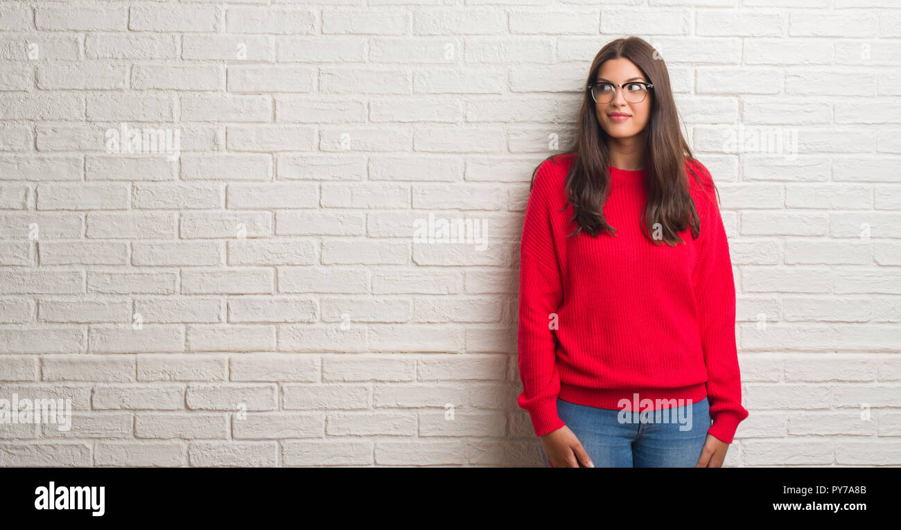 Young brunette woman standing over white brick wall smiling looking ...