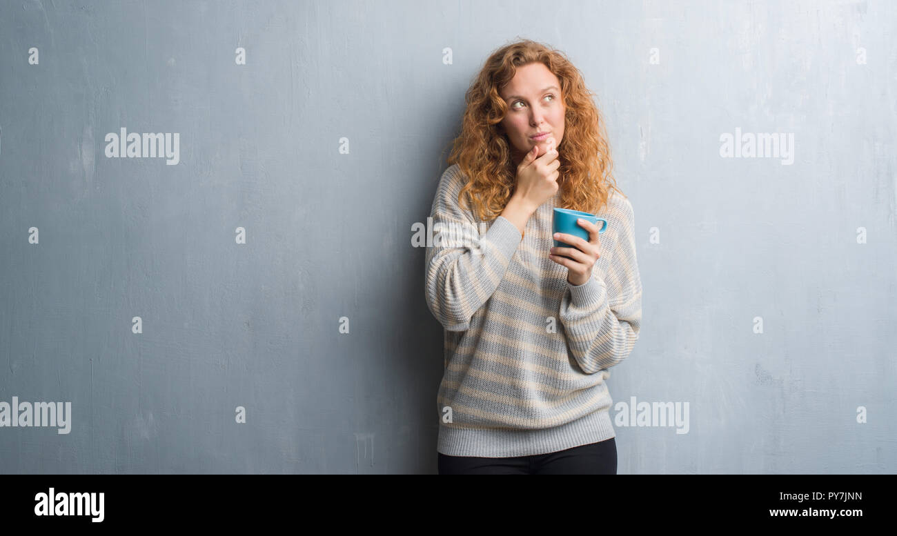 Young redhead woman over grey grunge wall drinking a cup of coffee ...