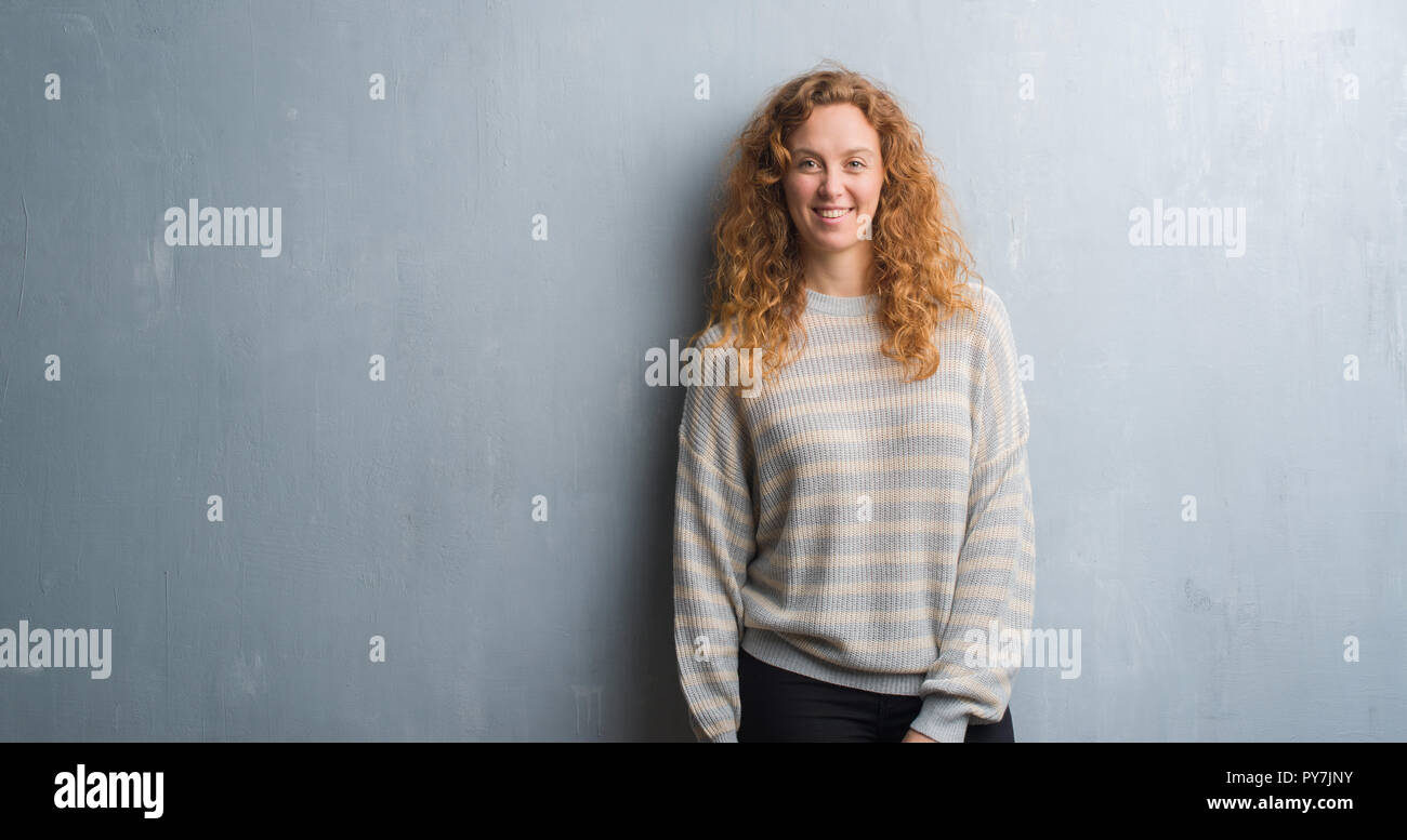 Young redhead woman over grey grunge wall with a happy and cool smile ...