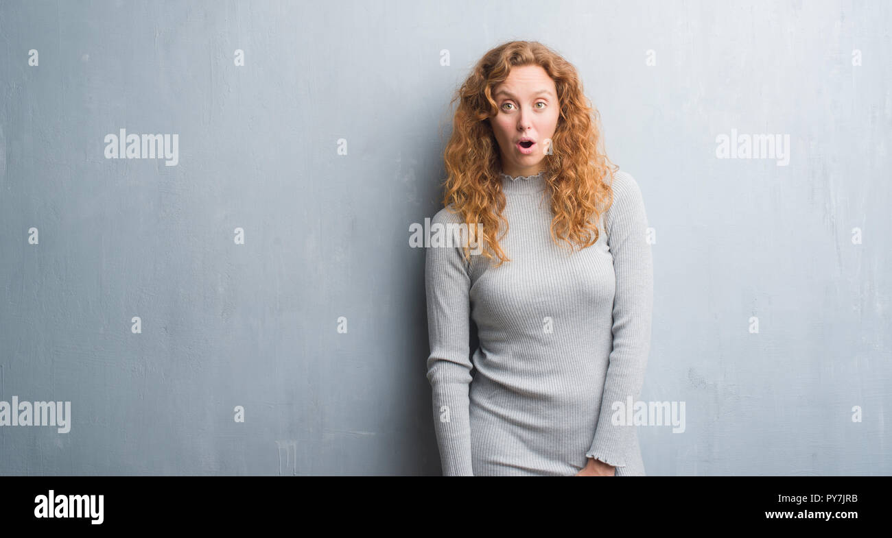Young redhead woman over grey grunge wall scared in shock with a ...