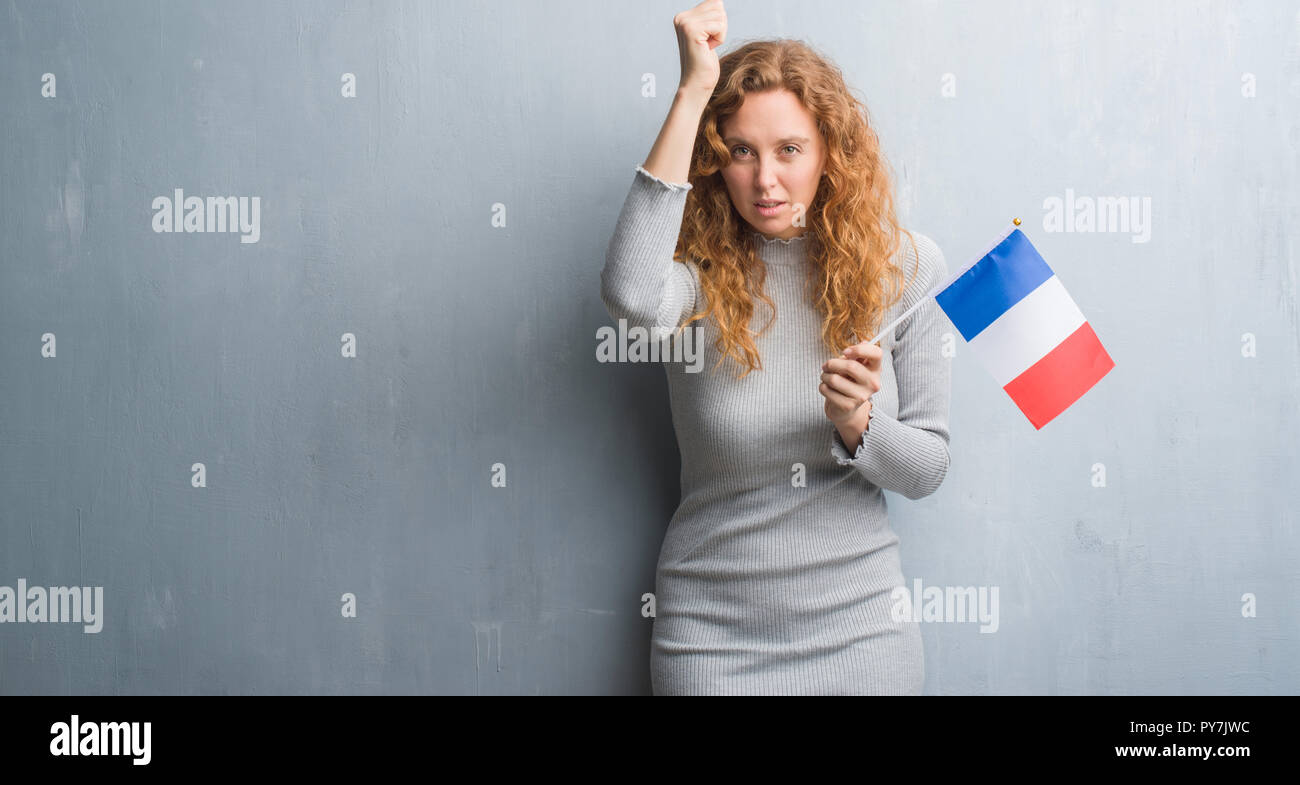 Young redhead woman over grey grunge wall holding flag of France ...