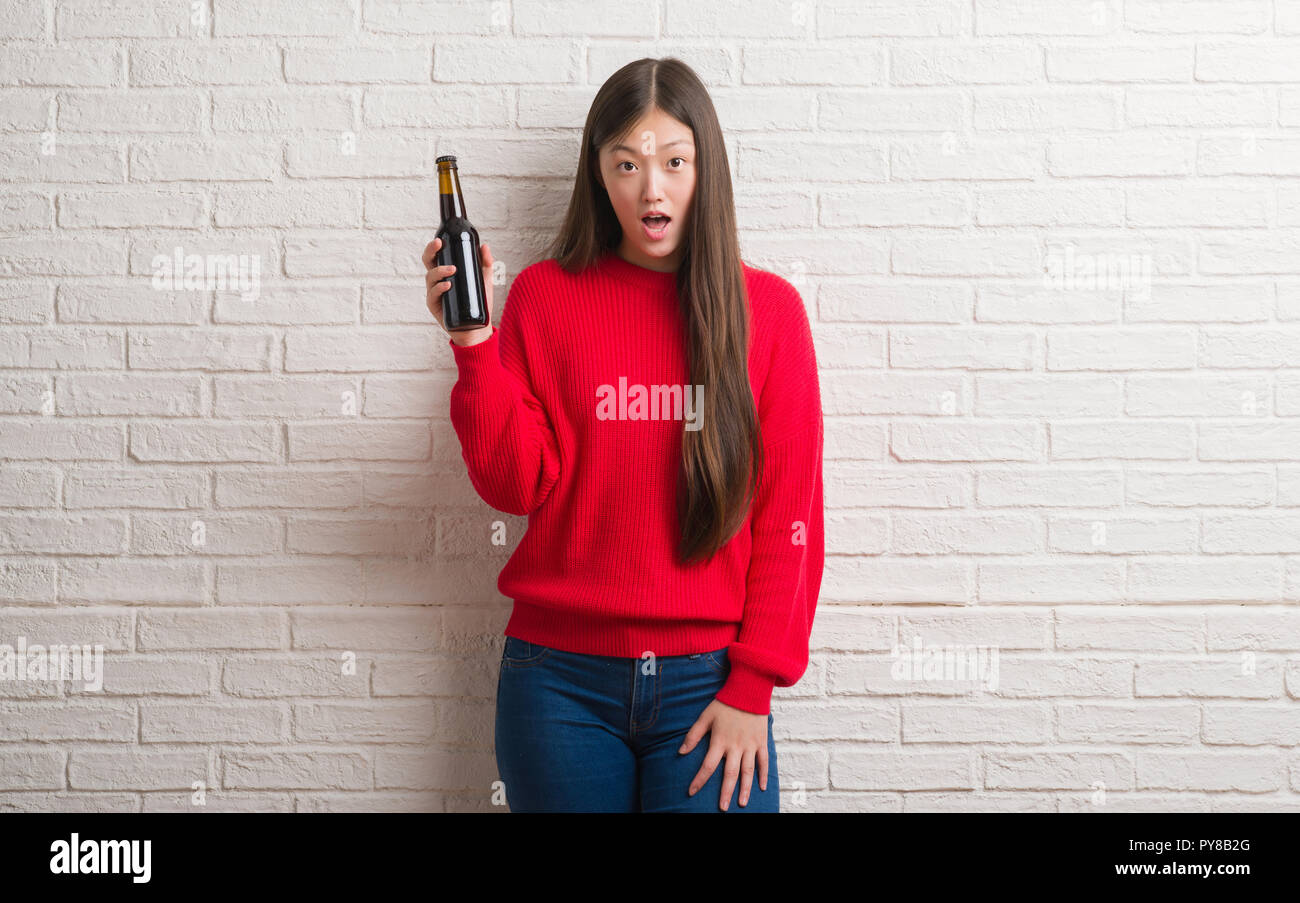 Young Chinese woman over brick wall drinking beer scared in shock with ...