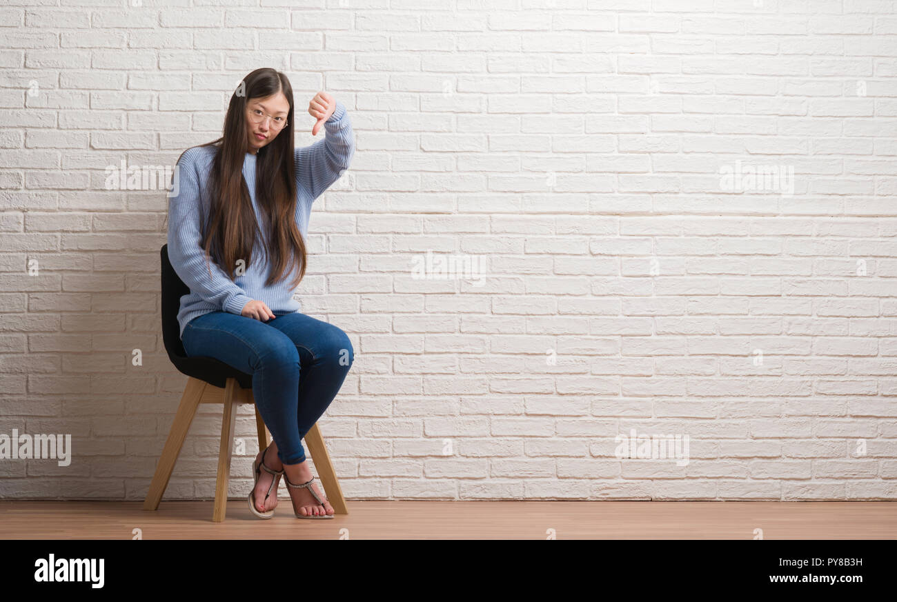Young Chinese woman sitting on chair over brick wall with angry face ...