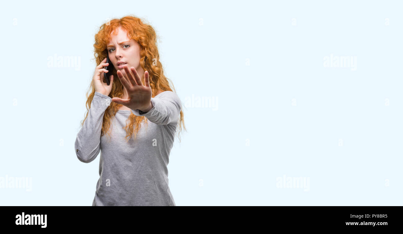 Young redhead woman talking on the phone with open hand doing stop sign ...