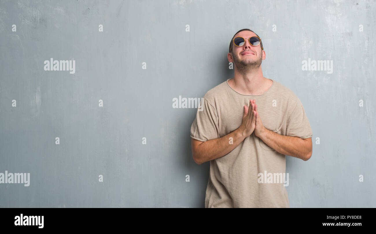 Young caucasian man over grey grunge wall wearing sunglasses begging ...