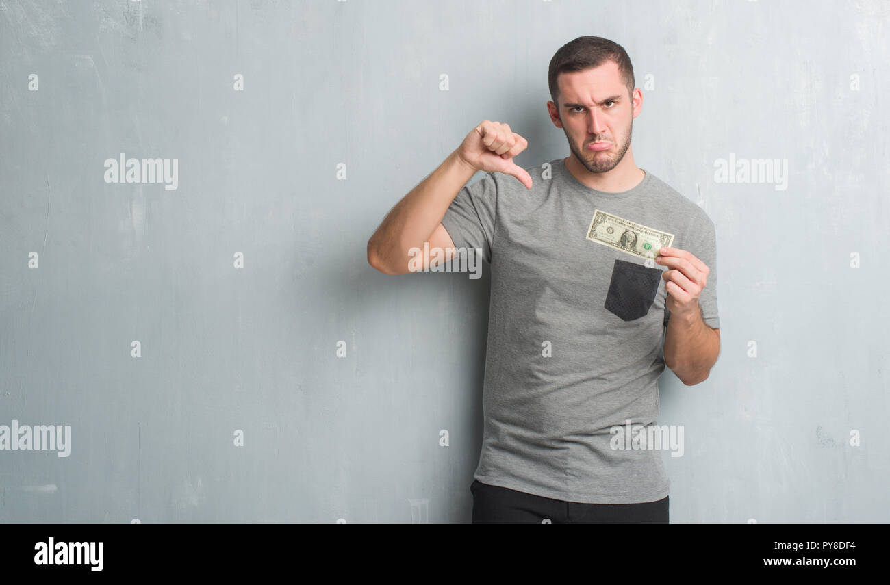Young caucasian man over grey grunge wall showing one dollar with angry ...