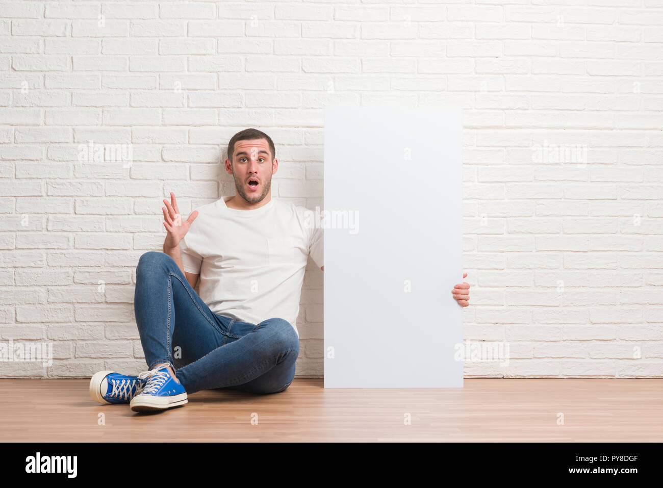 Young caucasian man sitting over white brick wall holding bottle of ...