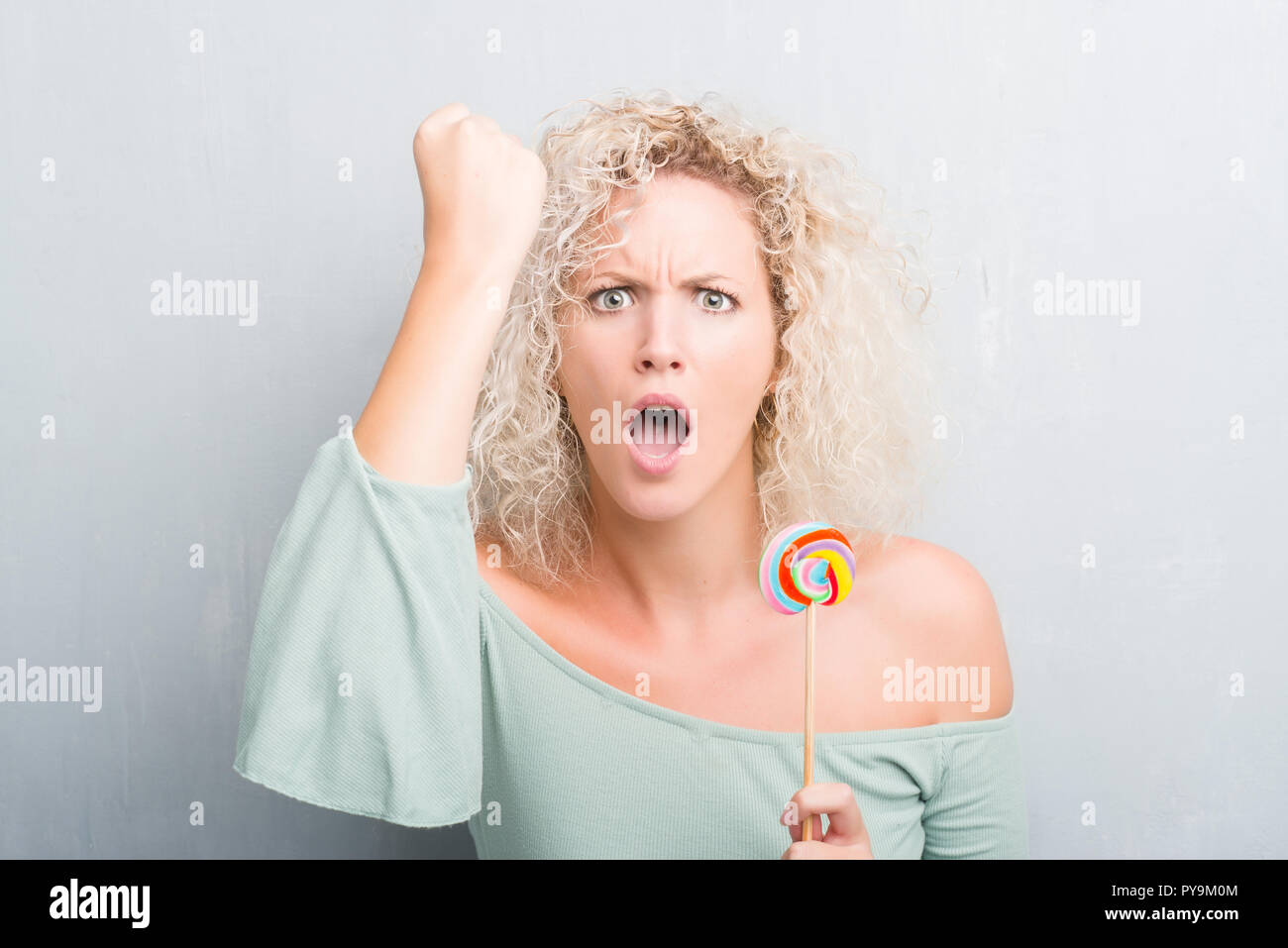 Young blonde woman over grunge grey wall eating candy lollipop annoyed ...