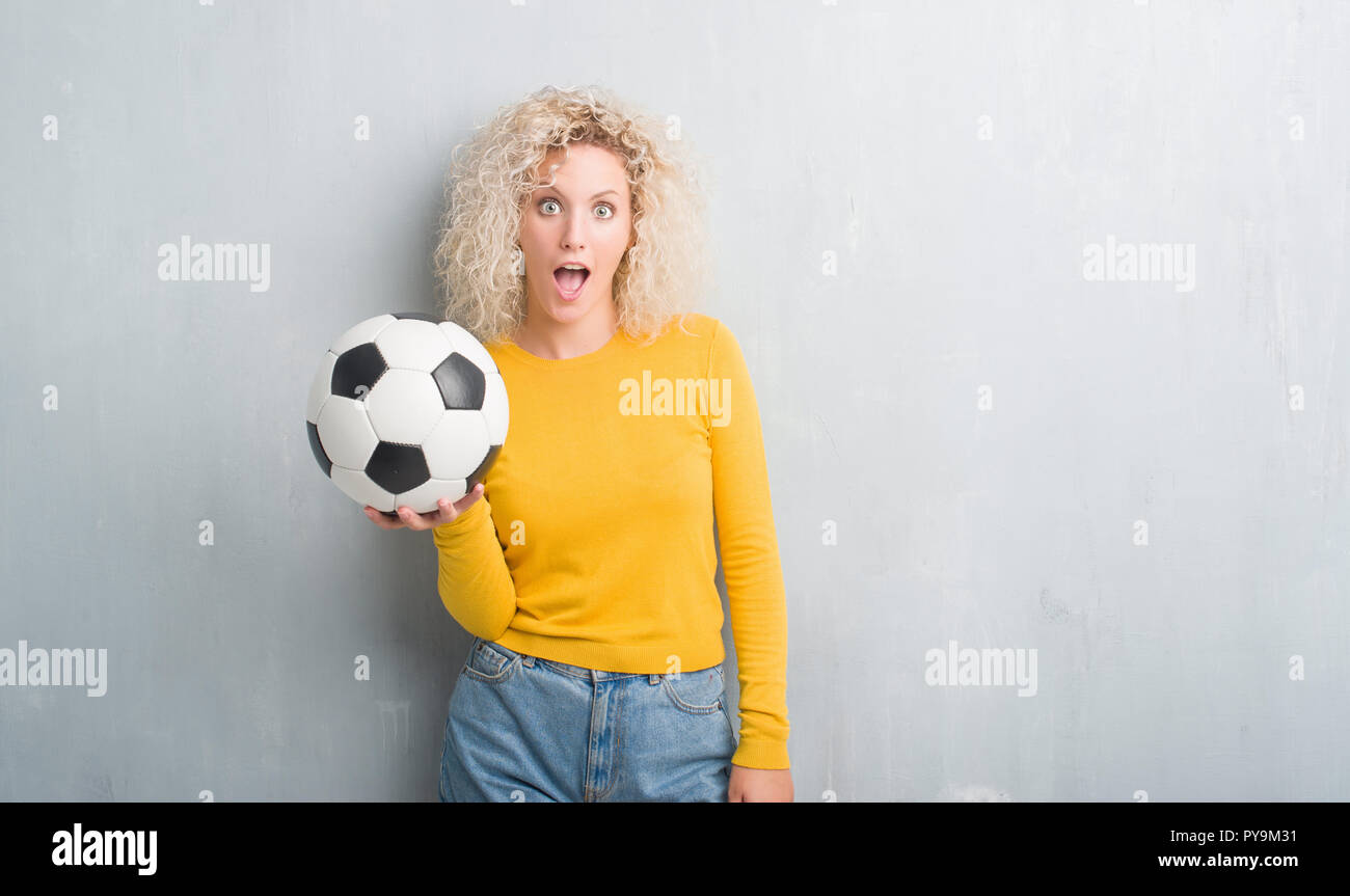 Young blonde woman over grunge grey background holding soccer football ...