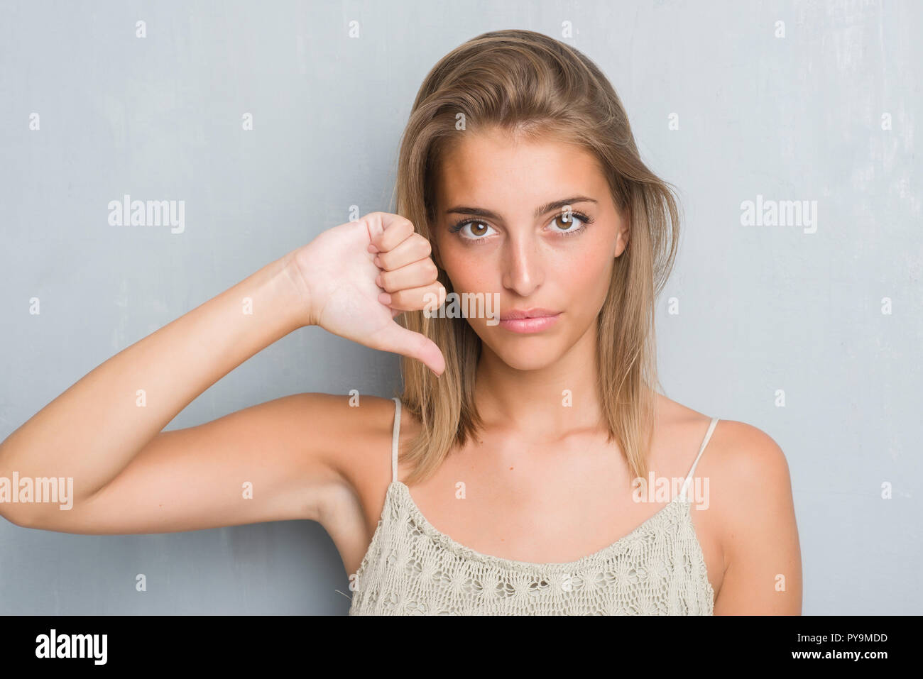 Beautiful young woman standing over grunge grey wall with angry face ...