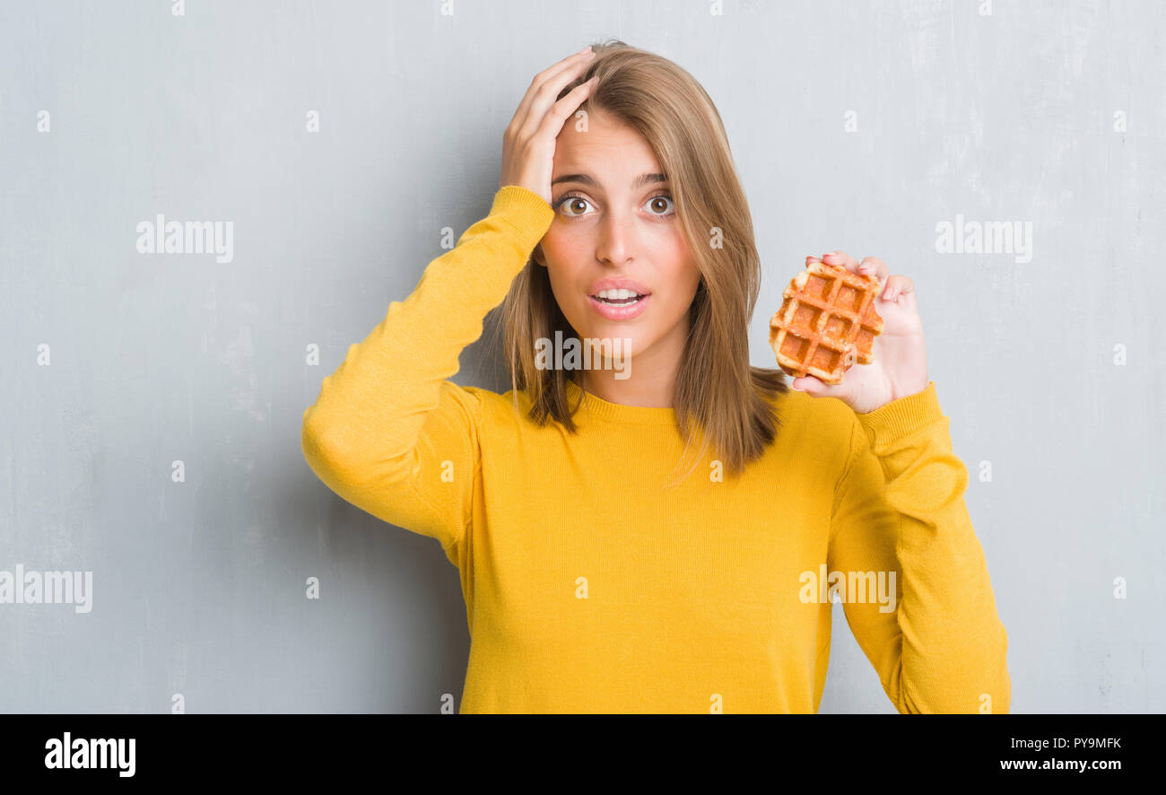 Beautiful young woman over grunge grey wall eating waffle stressed with ...