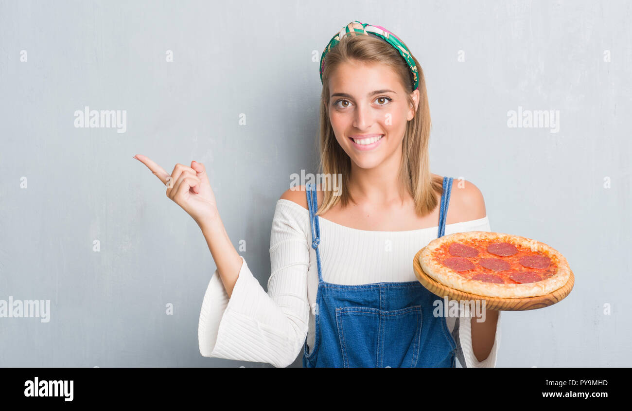 Beautiful young woman over grunge grey wall eating pepperoni pizza very ...
