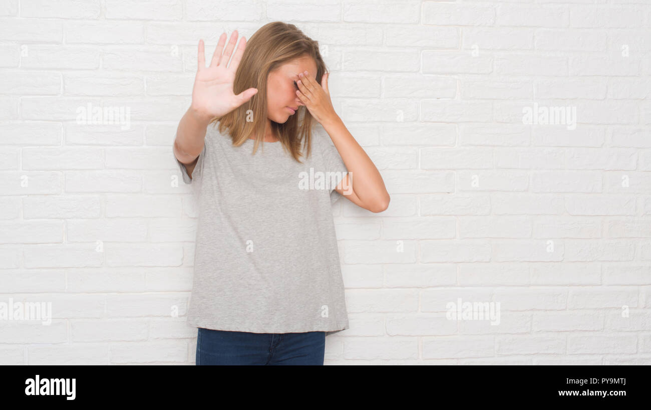 Beautiful young woman over white brick wall covering eyes with hands ...