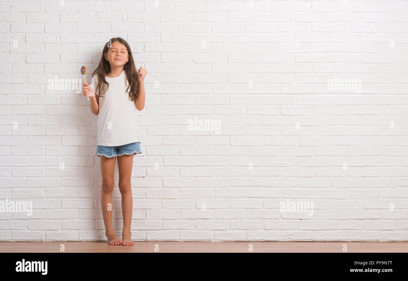 Young hispanic kid over white brick wall eating lollipop candy at home ...