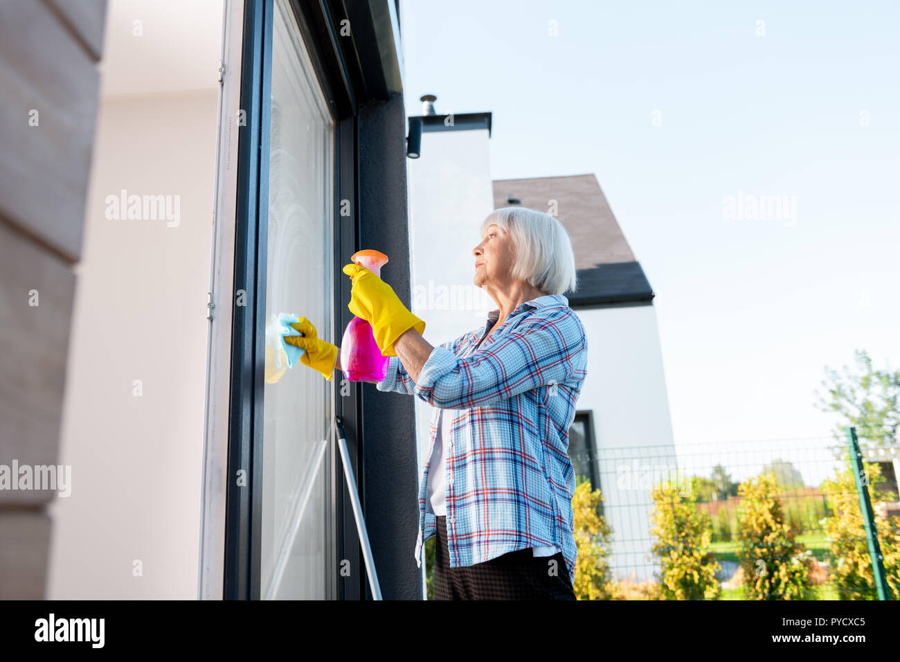 Elderly housewife wearing blue squared shirt washing windows outside ...