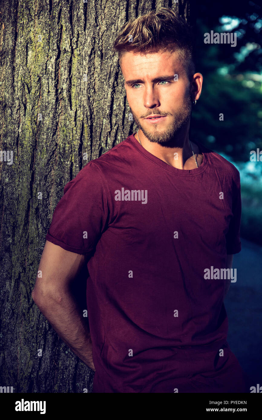 Sad, worried blond young man leaning against tree, outside in nature ...