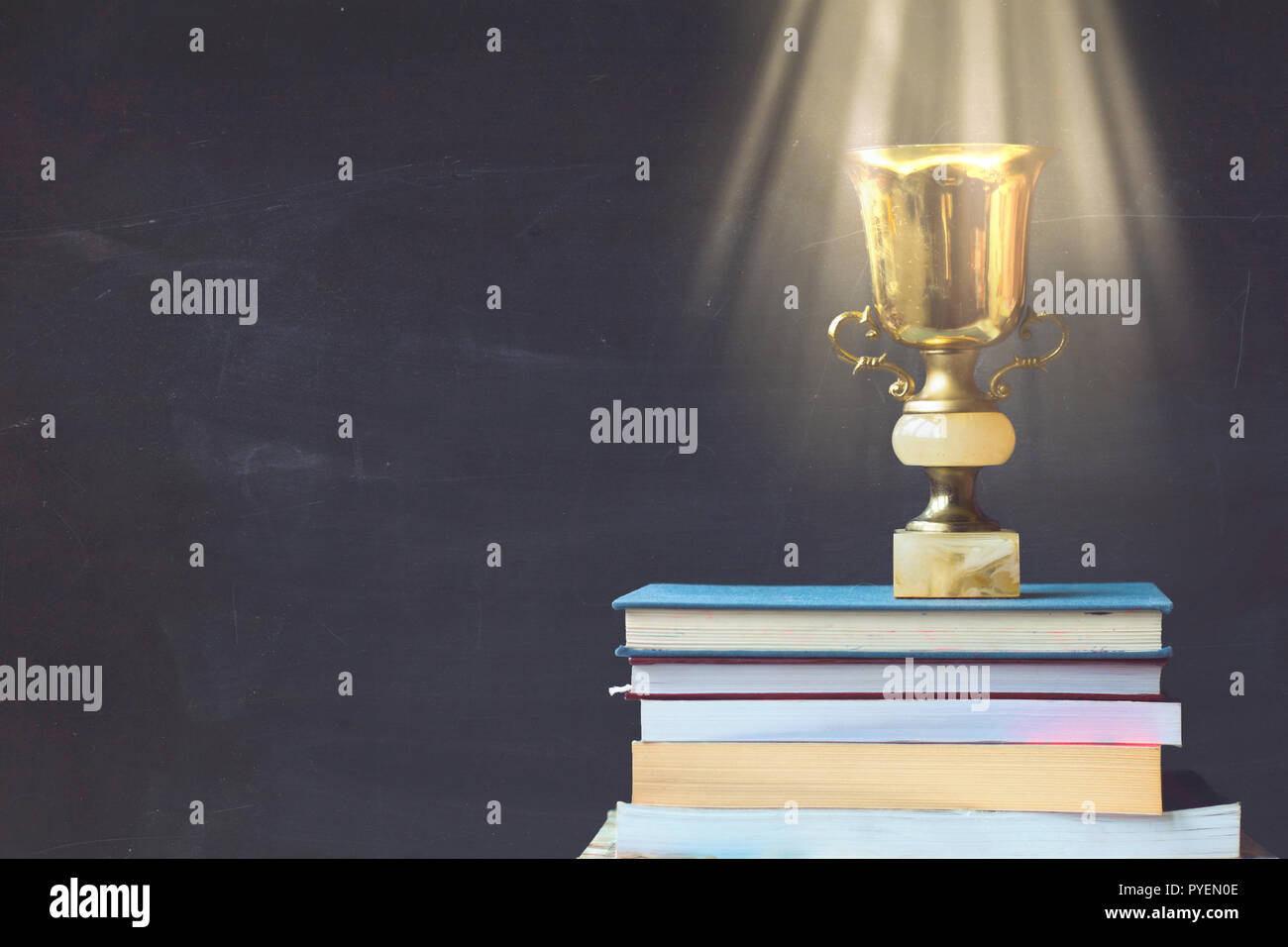 Golden trophy on pile of books, against blackboard, with sun rays over ...