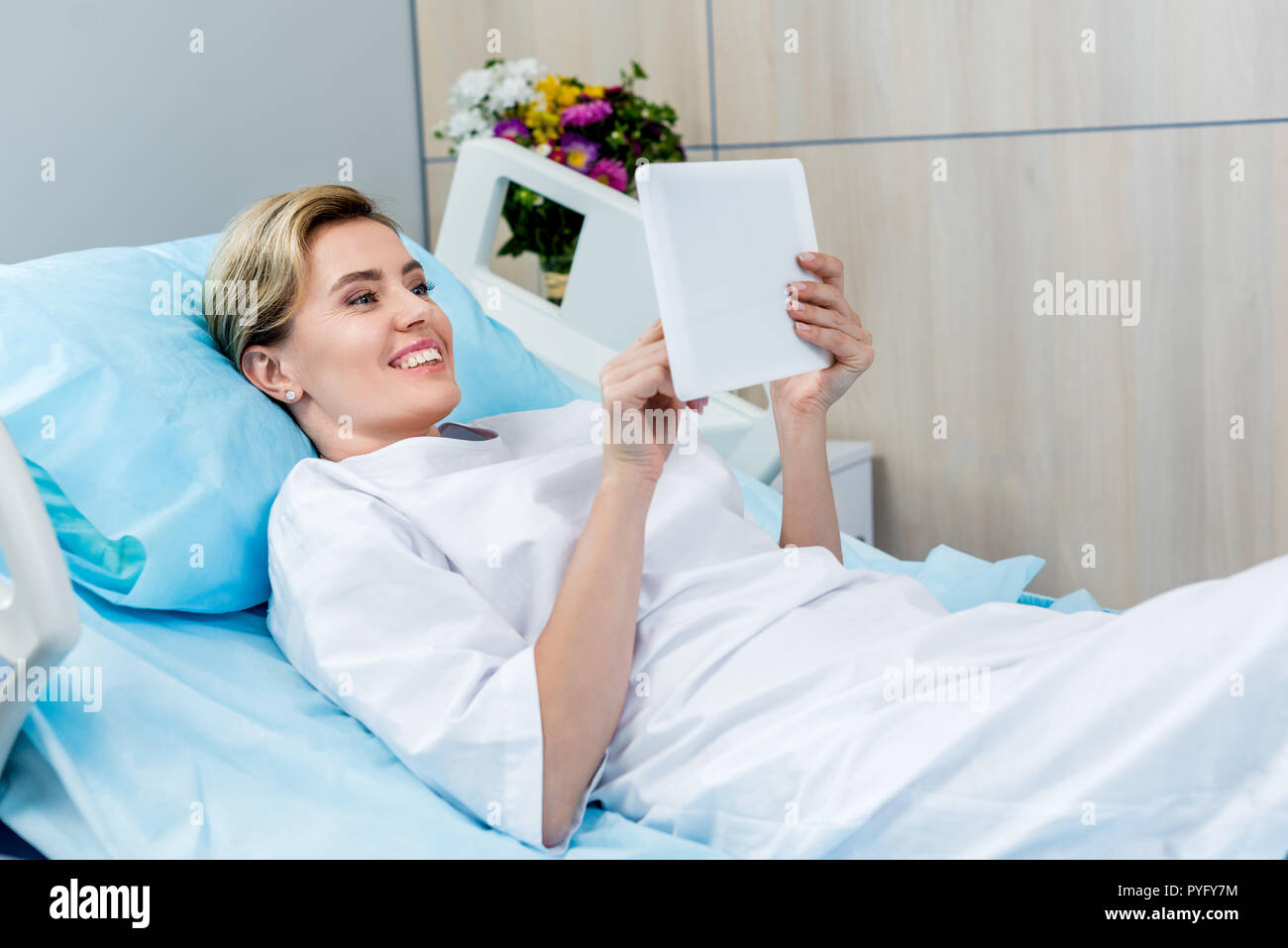 smiling adult female patient using digital tablet on bed in hospital ...