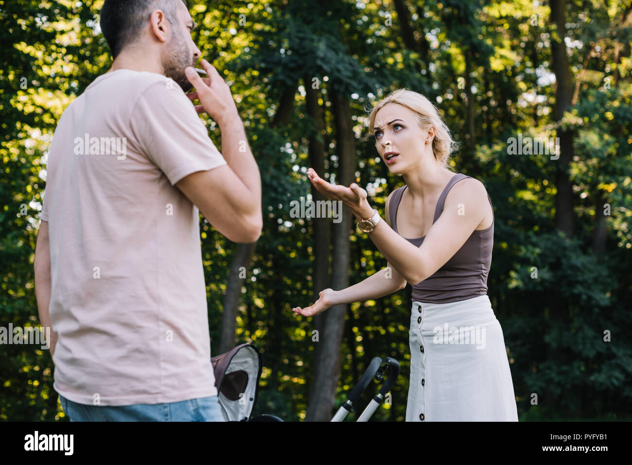 father smoking cigarette near baby carriage in park, mother gesturing ...