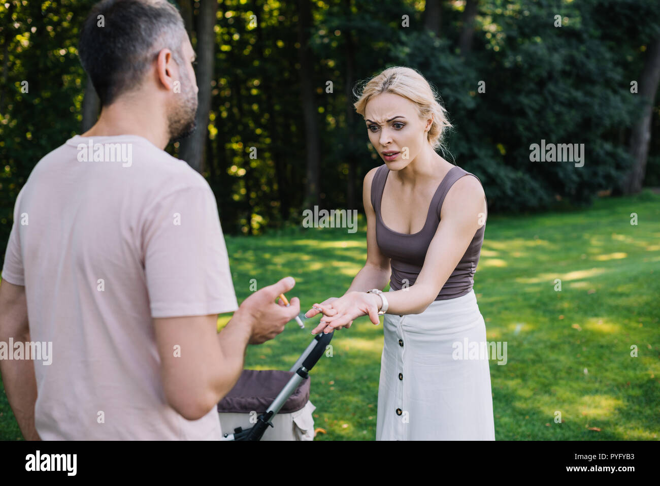 father smoking cigarette near baby carriage in park, mother pointing on ...